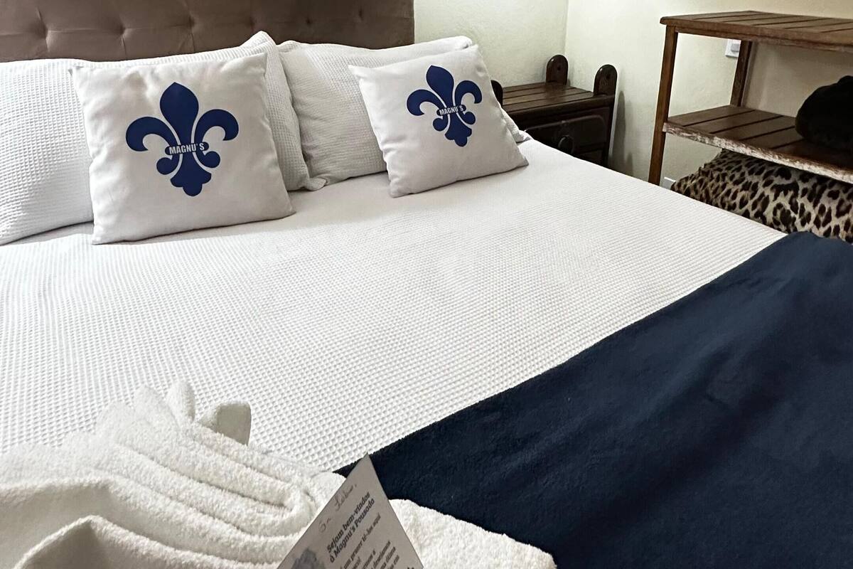 Cama bem arrumada com roupa de cama branca e duas almofadas com o desenho de lis, uma mesa de cabeceira com abajur e uma pequena mesa com almofada estampada em leopardo.