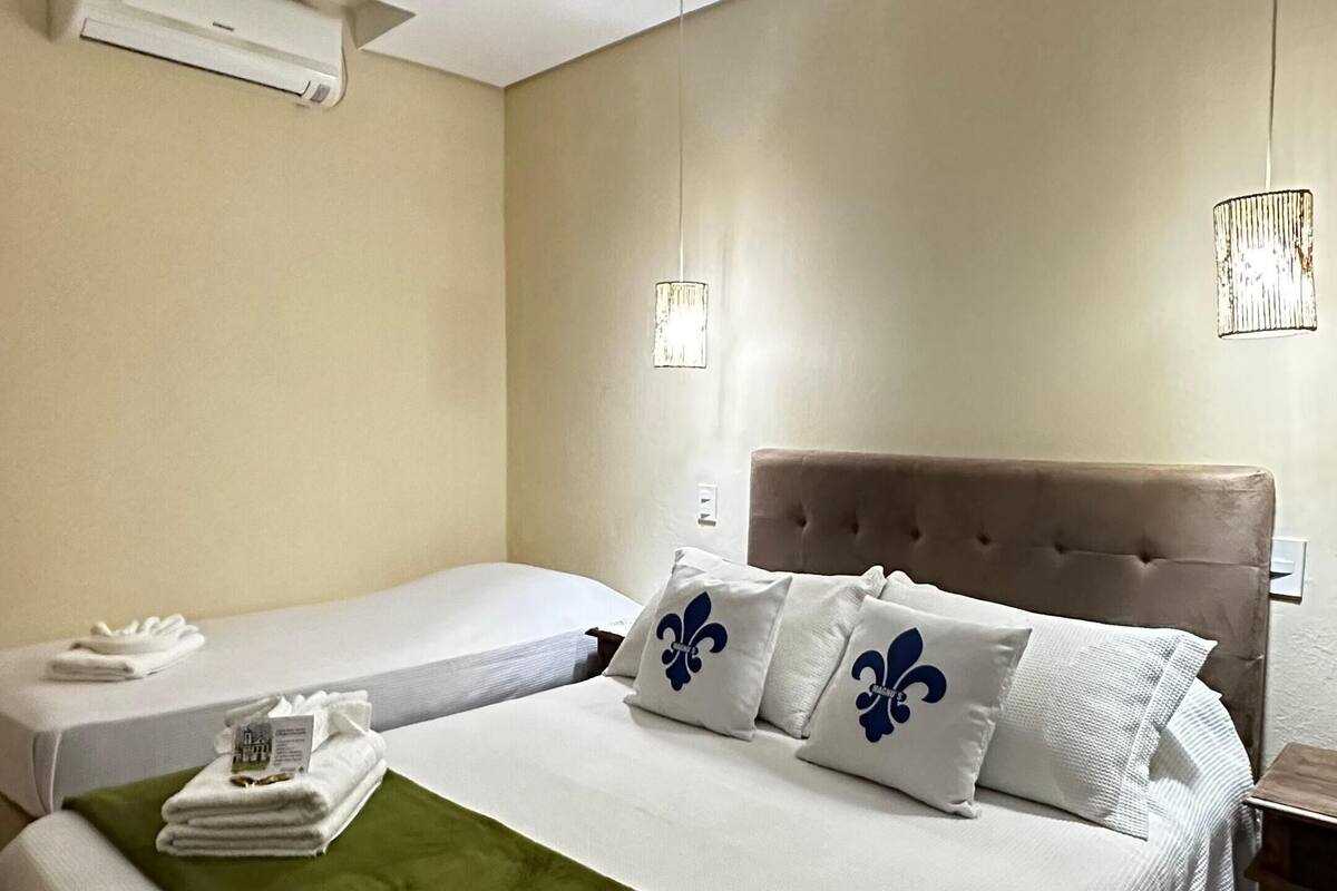 Quarto de hotel com cama, mesa de cabeceira, luminária e ar condicionado fixado na parede.