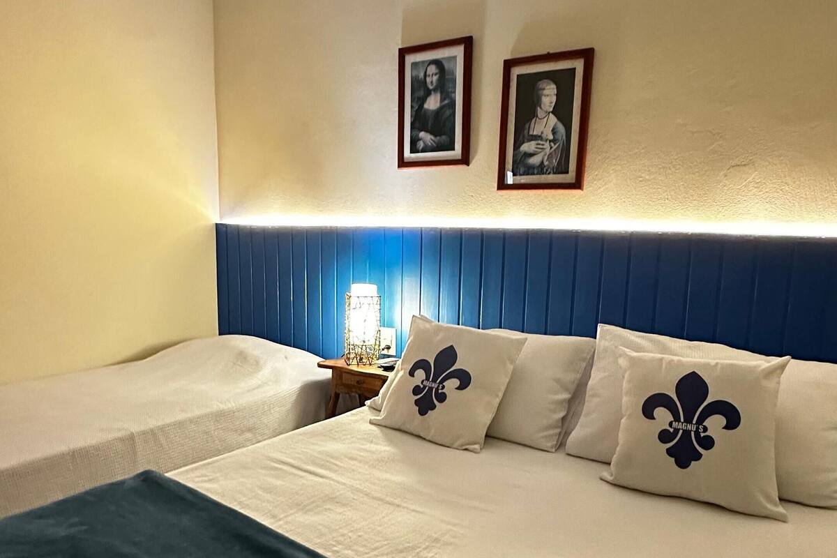 Quarto de hotel com cama, duas almofadas com desenhos de flores-de-lis, cabeceira azul e mesa de cabeceira com luminária.