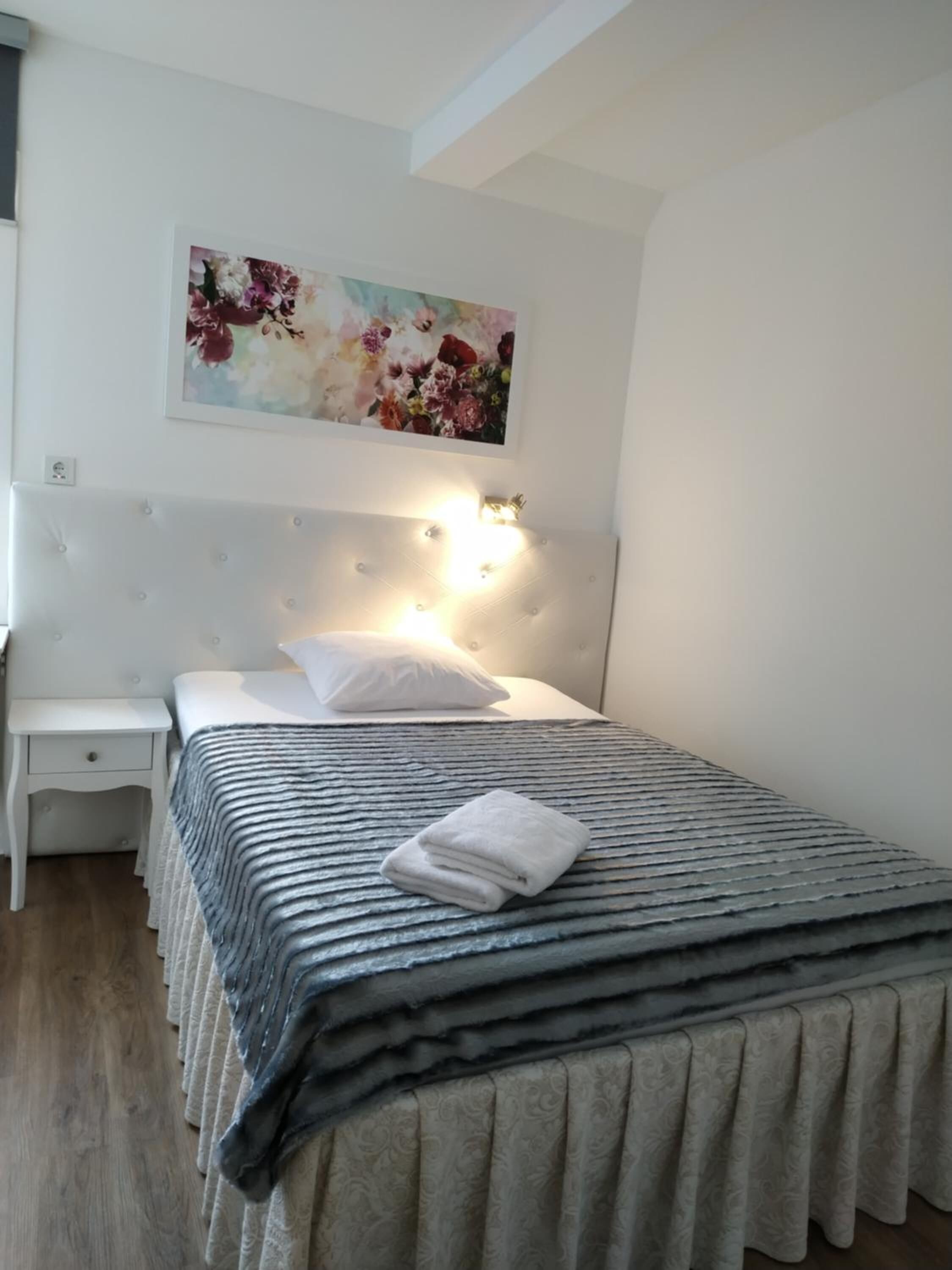 Uma cama bem arrumada com colcha listrada e uma toalha dobrada, uma mesinha de cabeceira com gaveta, e um candeeiro fixado na parede.
