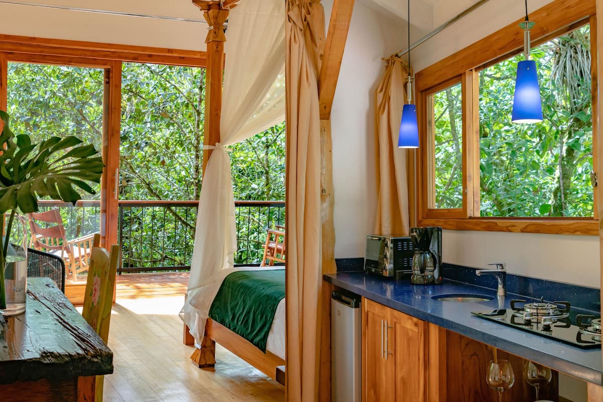 Quarto com cama, pequeno espaço de cozinha, mesa de jantar com cadeiras e uma janela grande com cortinas.