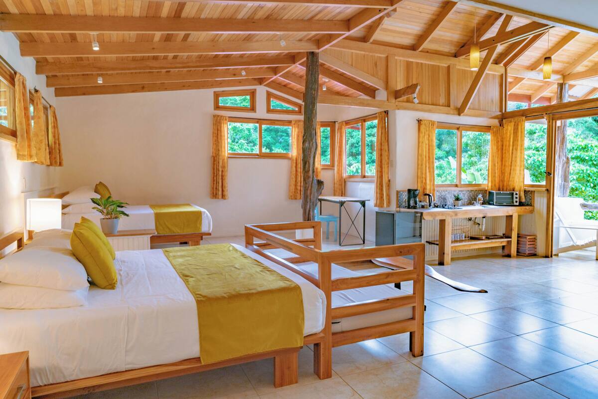 Quarto com duas camas de madeira, uma kitchenette pequena e grandes janelas com vista para o verde.