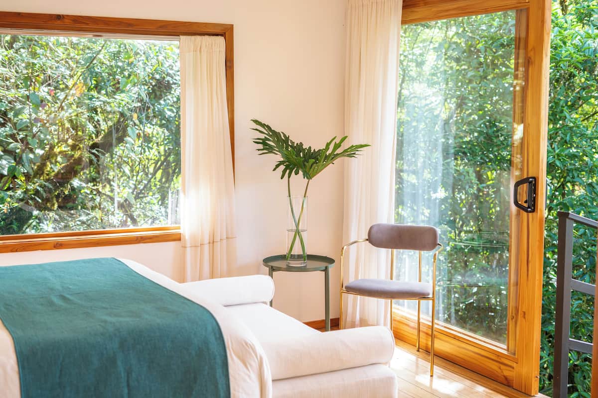 Quarto com cama, uma cadeira, uma mesa com planta e uma grande janela com cortinas.