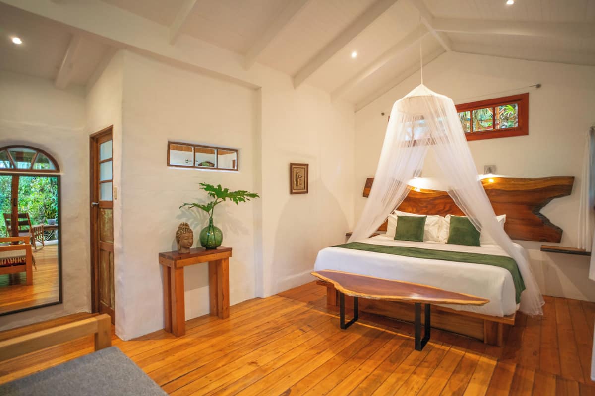 Quarto com cama de madeira, mesa de cabeceira, planta em vaso e vista para o exterior.