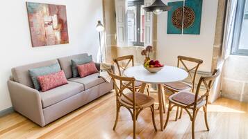 Uma sala de estar aconchegante com um sofá bege, cadeiras de madeira, uma mesa branca com uma cesta de frutas e um objeto decorativo na parede.