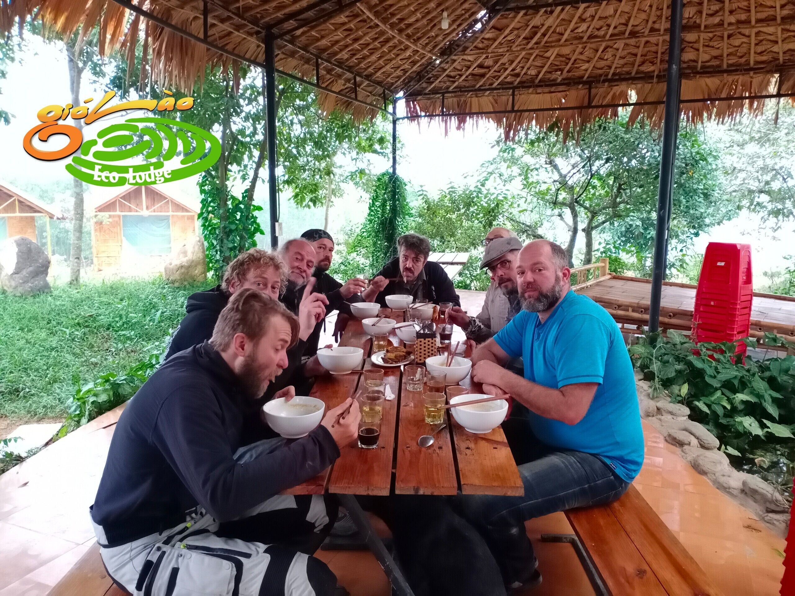 Um grupo de pessoas jantando em uma mesa de madeira com tigelas e copos, sob um telhado de palha, cercado por vegetação.