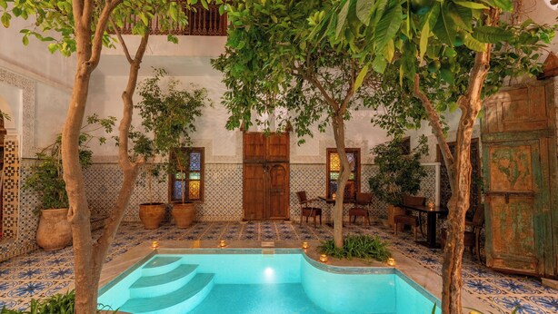 Uma área de piscina com piso em azulejo, móveis de madeira e plantas em vasos.