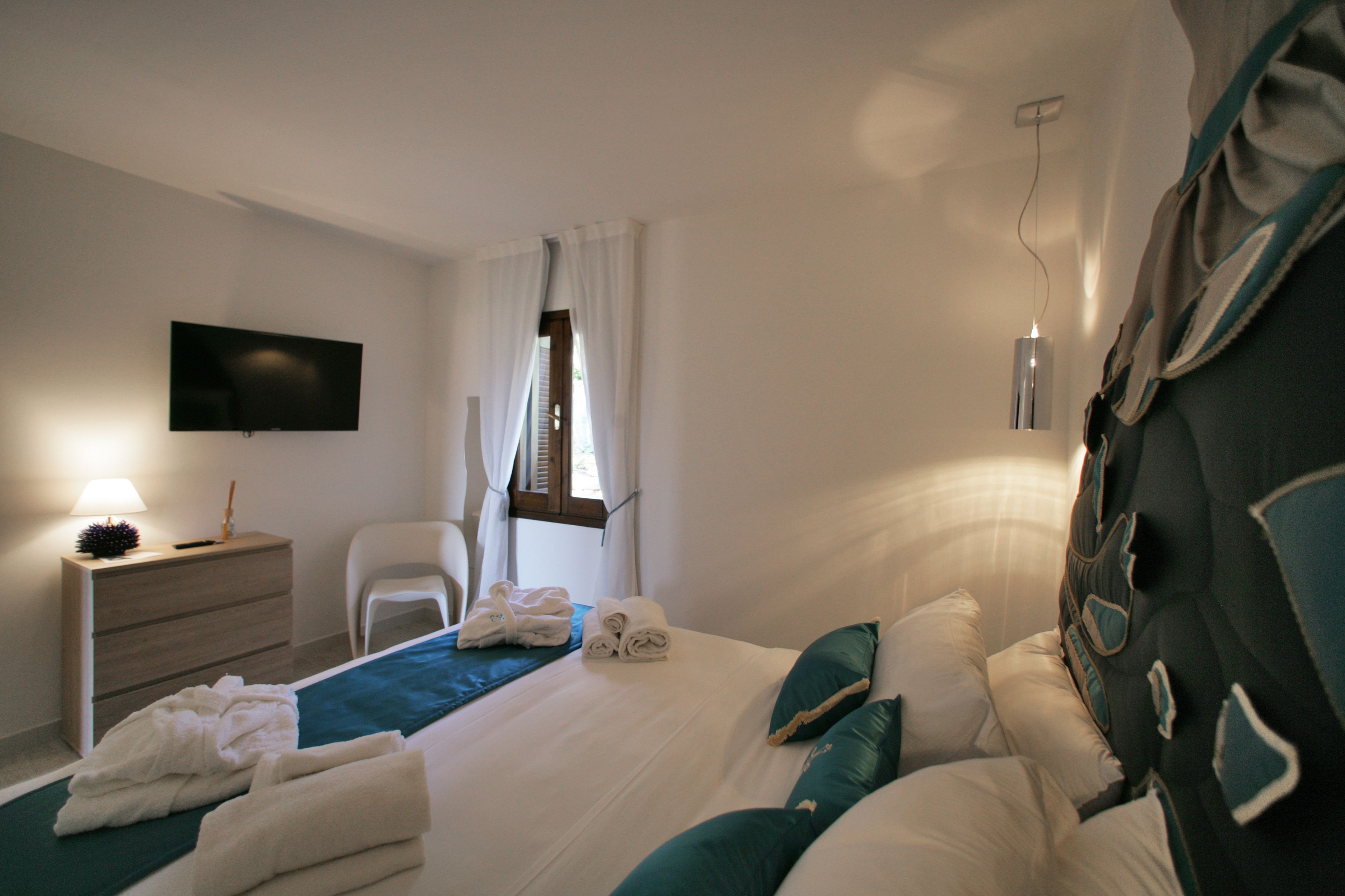 Quarto de hotel com cama, escrivaninha, televisão e janela com cortinas.