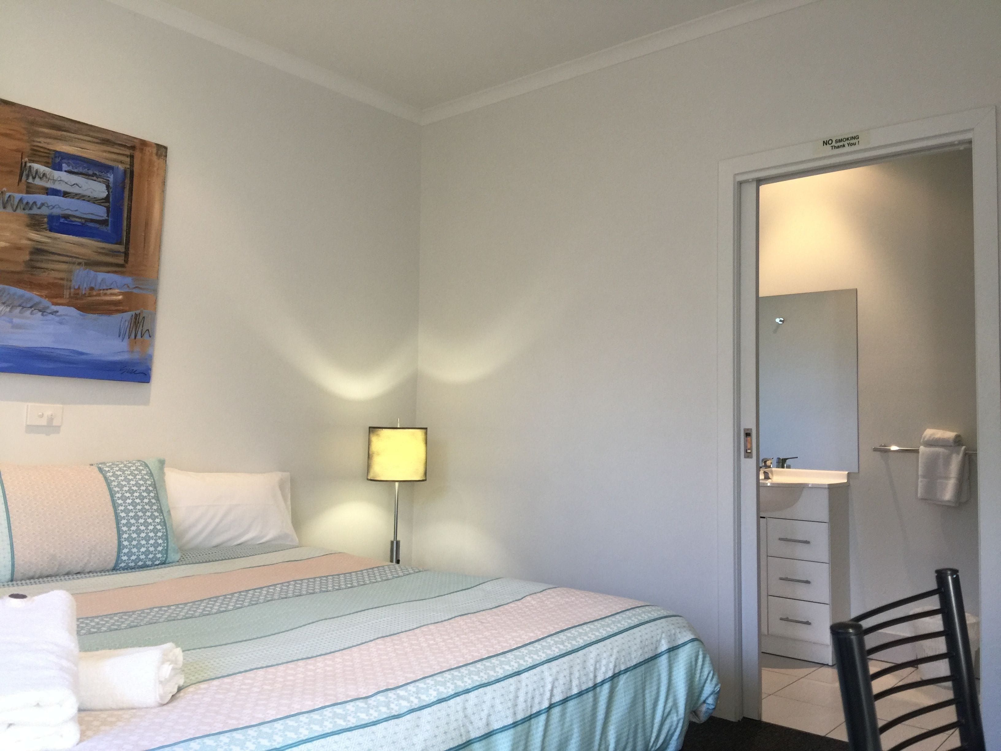 Quarto de hotel com cama, mesa de cabeceira, luminária, porta e banheiro visível através da porta.