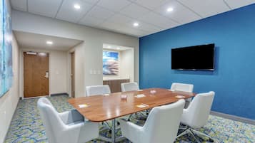 Sala de conferências com mesa de madeira, cadeiras brancas, parede azul decorativa e TV fixada.