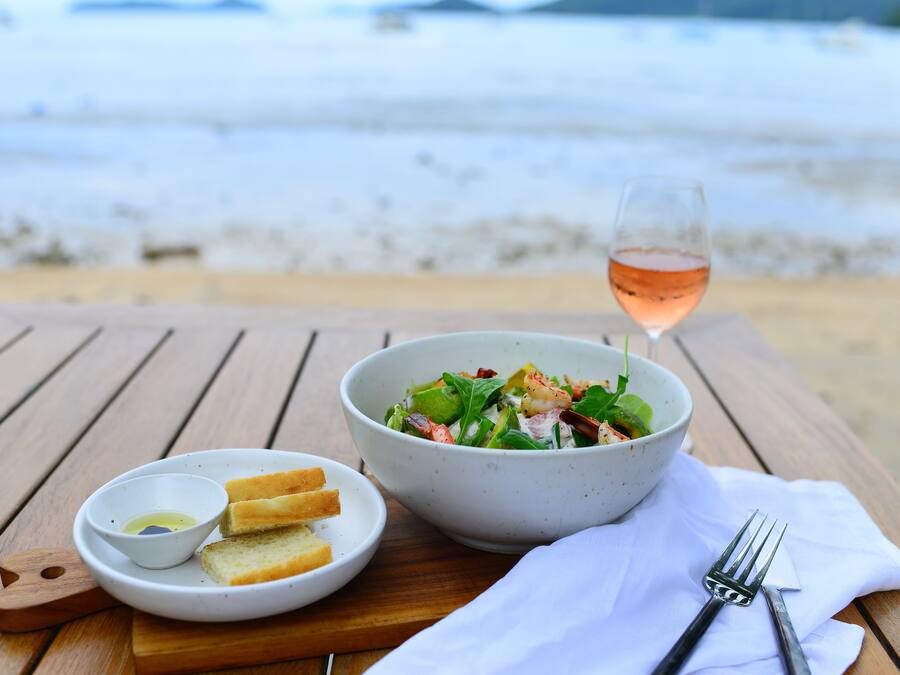 Uma mesa de madeira com uma salada numa tigela, um copo de vinho e um pequeno recipiente com molho.