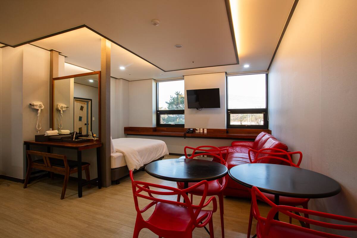 Quarto de hotel compacto com cama, escrivaninha com espelho, televisão fixada na parede e cadeiras vermelhas ao redor de uma mesa pequena.