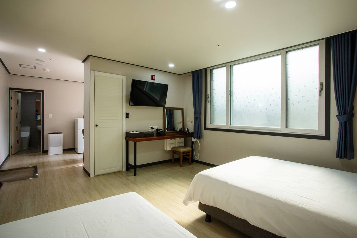 Quarto de hotel com duas camas, uma escrivaninha com espelho, televisão e janela com cortinas.