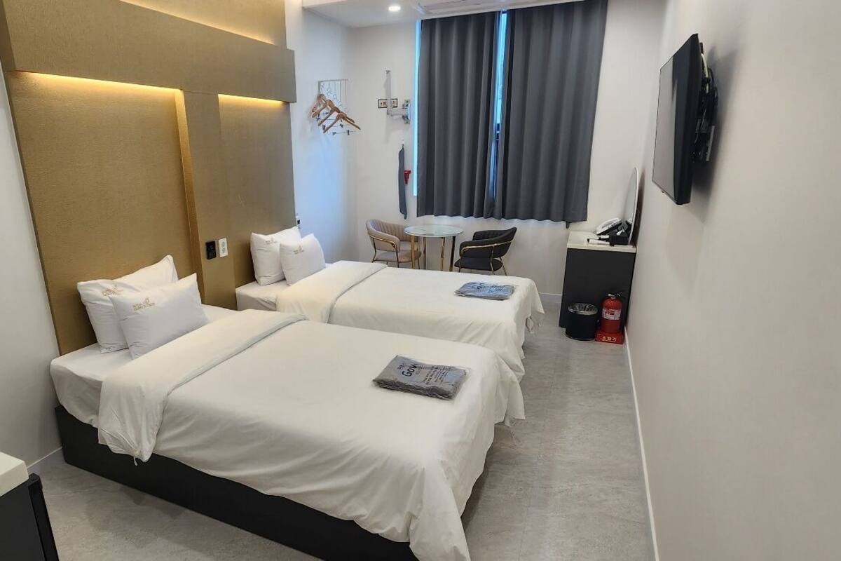 Quarto de hotel com duas camas, uma mesa pequena, cadeiras, uma televisão e uma janela com cortinas.