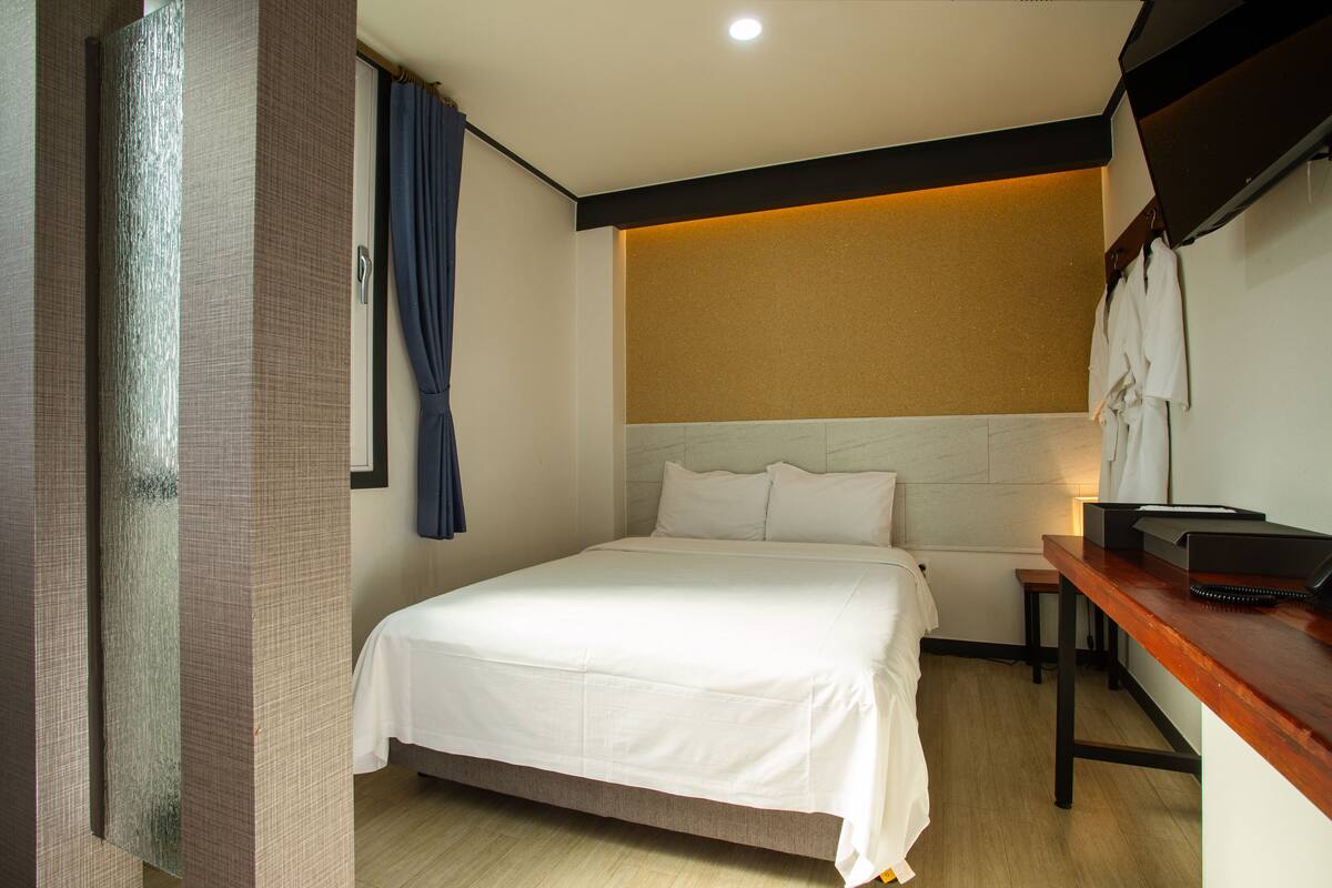 Quarto de hotel com uma cama, uma mesinha e uma televisão fixada na parede.