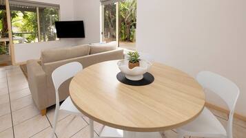 Uma sala de estar moderna com um sofá bege, uma mesa de jantar redonda de madeira com cadeiras brancas, uma TV de tela plana na parede e grandes janelas com vista para vegetação.