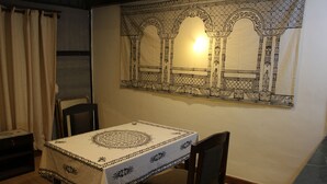 Uma área de refeições com uma mesa coberta por uma toalha estampada, duas cadeiras e uma tapeçaria na parede retratando um desenho arquitetônico.