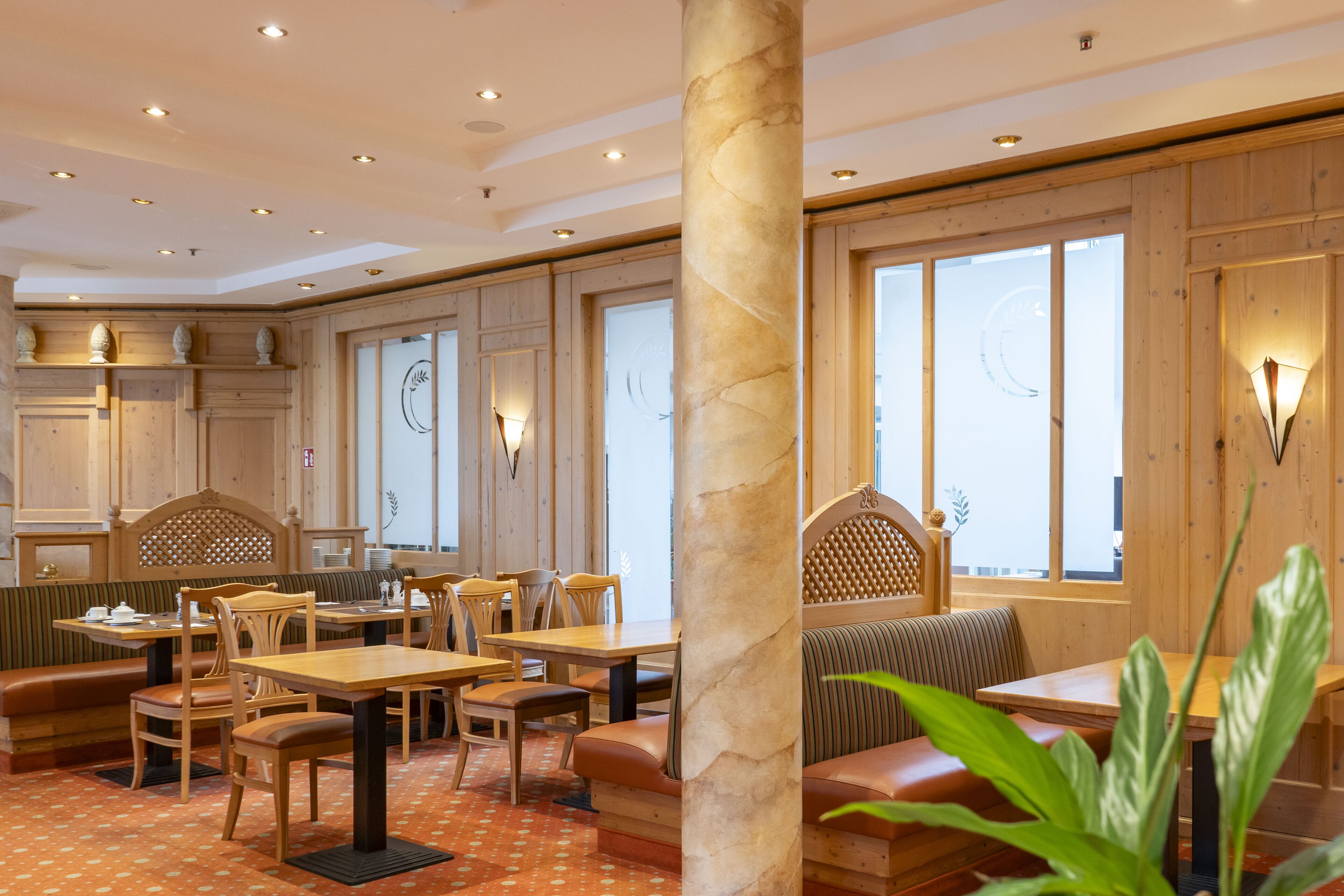 Um restaurante com painéis de madeira, diversas áreas de estar incluindo cabines e mesas, e grandes janelas com painéis de vidro decorativos.