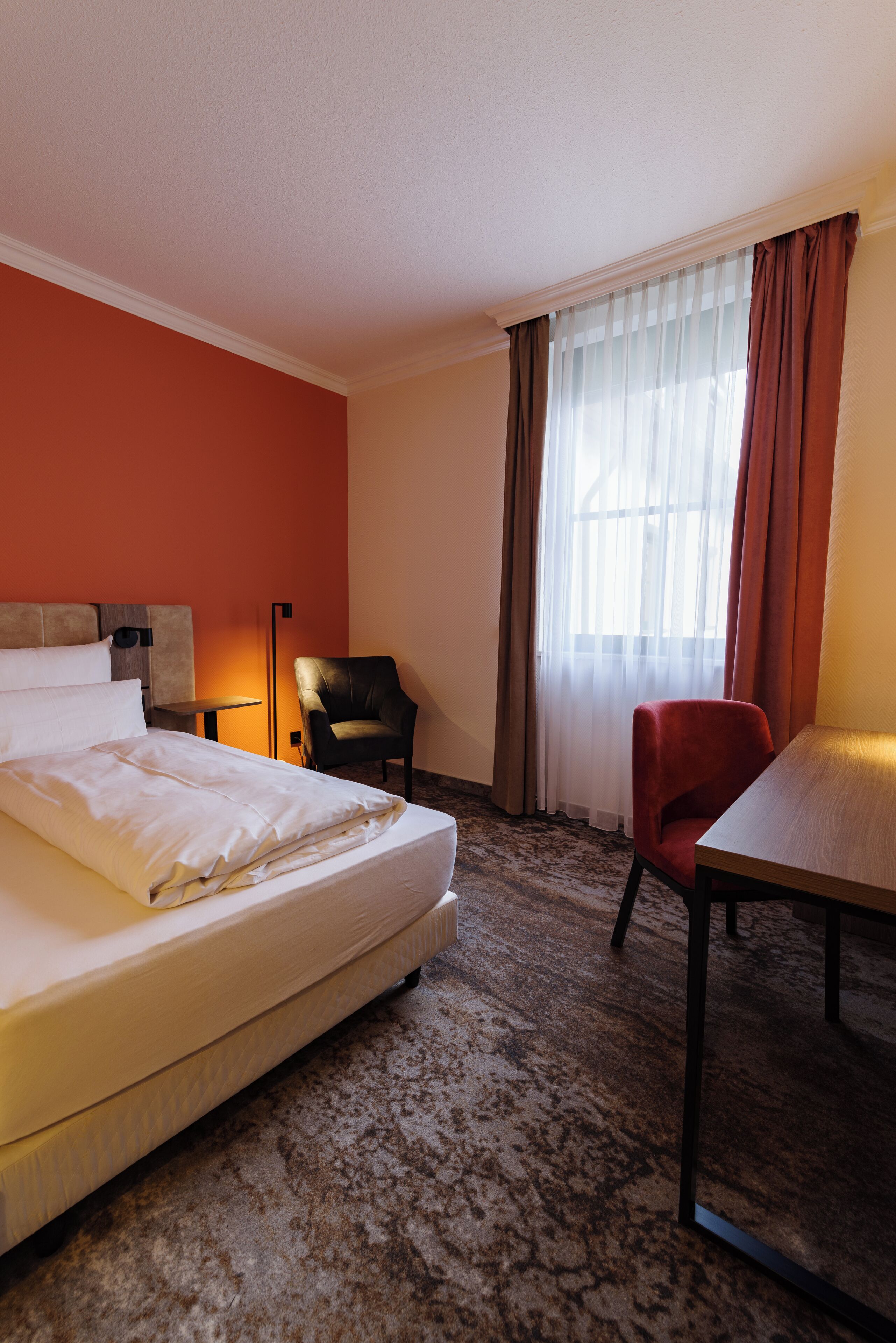 Quarto de hotel com cama, escrivaninha, cadeira e luminária.