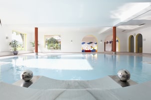 Uma grande piscina interior com água cristalina, rodeada por paredes brancas e algumas janelas.