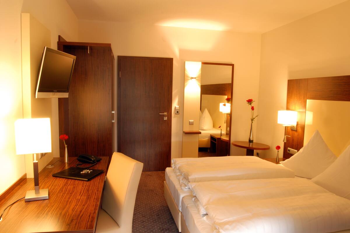 Quarto de hotel com cama, mesas de cabeceira, escrivaninha com luminária, televisão e espelho.