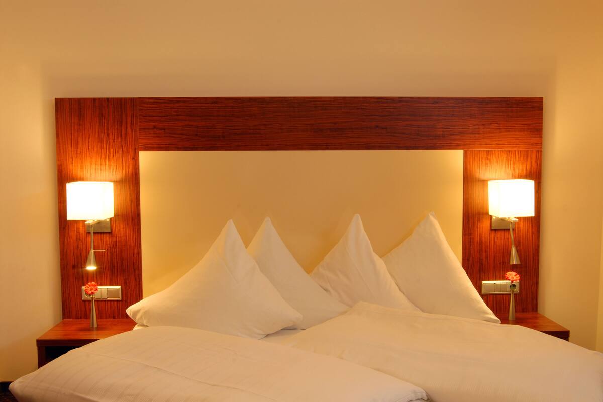 Quarto de hotel com cabeceira de madeira, duas luminárias de mesa e cama com lençóis brancos.