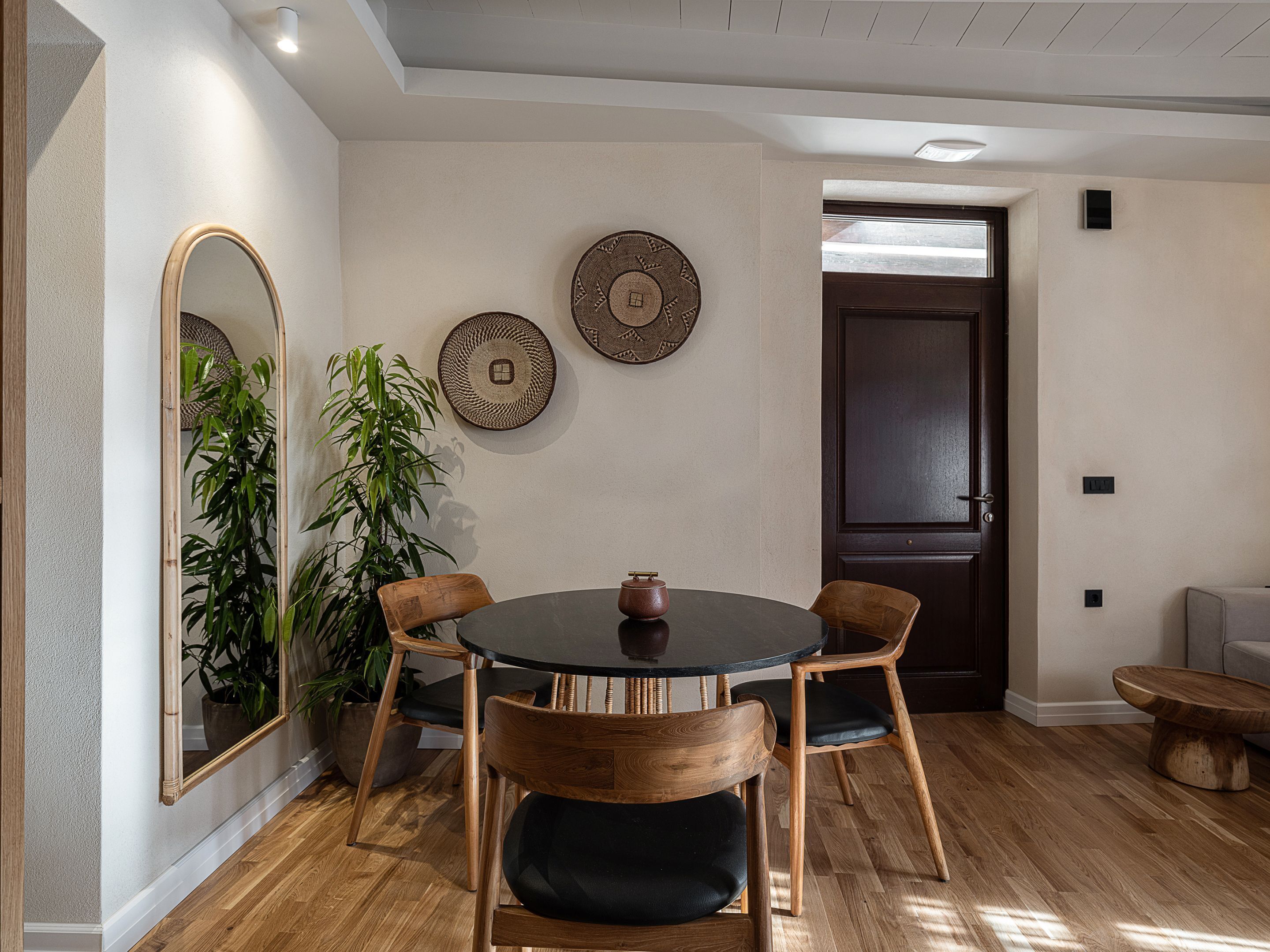 Uma sala de jantar com uma mesa redonda, quatro cadeiras de madeira, dois pratos decorativos pendurados na parede, um espelho e um vaso com planta.