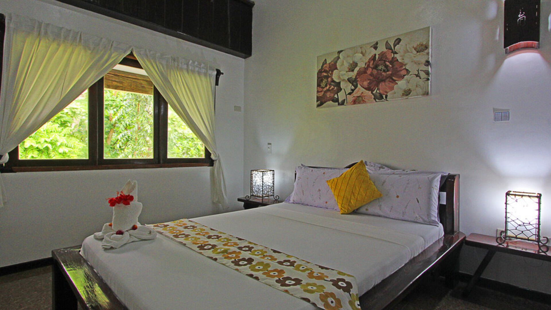 Cama bem arrumada com colcha florida, um travesseiro amarelo e uma toalha branca ao lado. Há um quadro de flores na parede acima da cama.