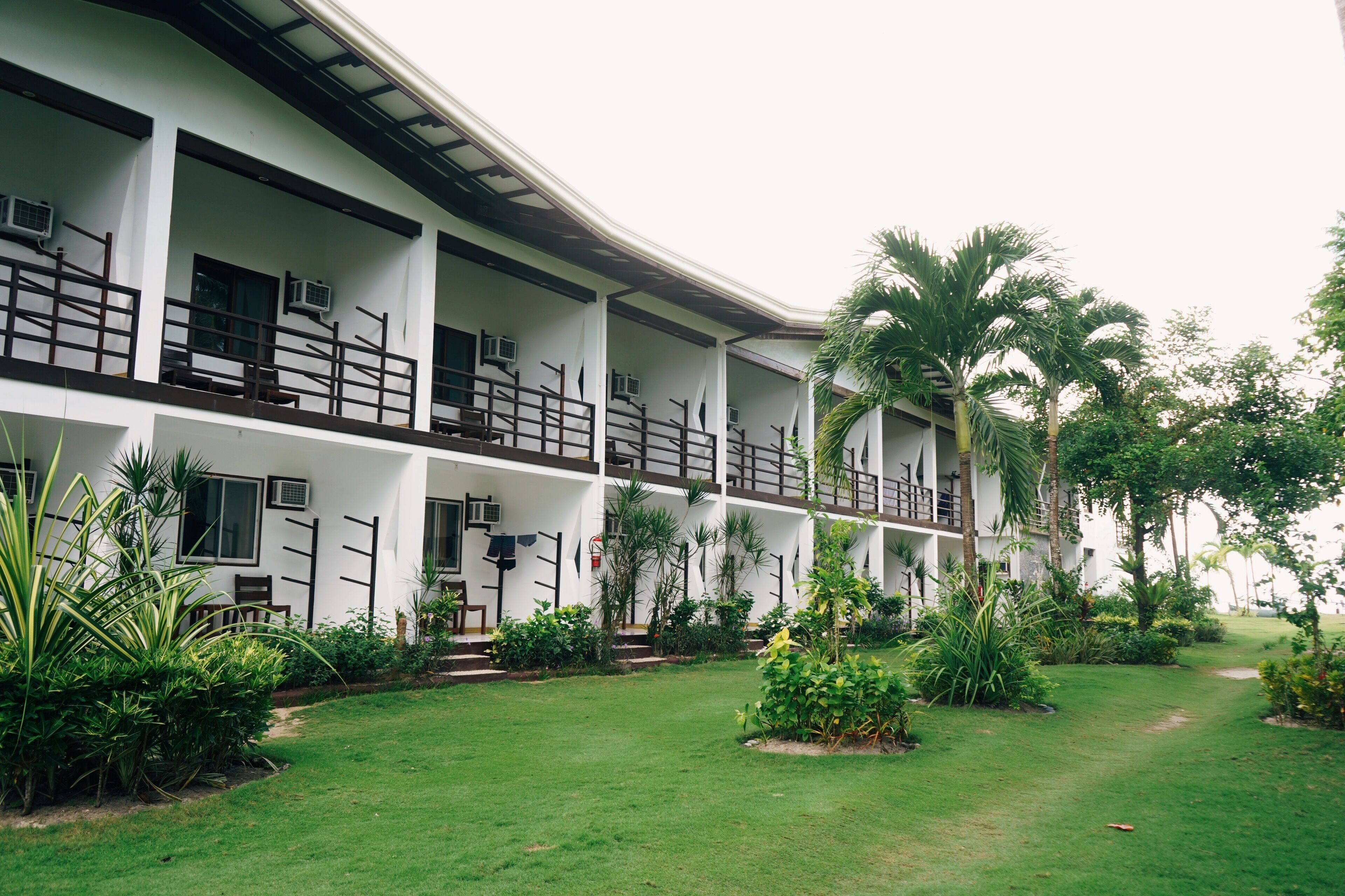 Hotel de vários andares com varandas, aparelhos de ar condicionado e um jardim bem cuidado com diversas plantas e uma palmeira.