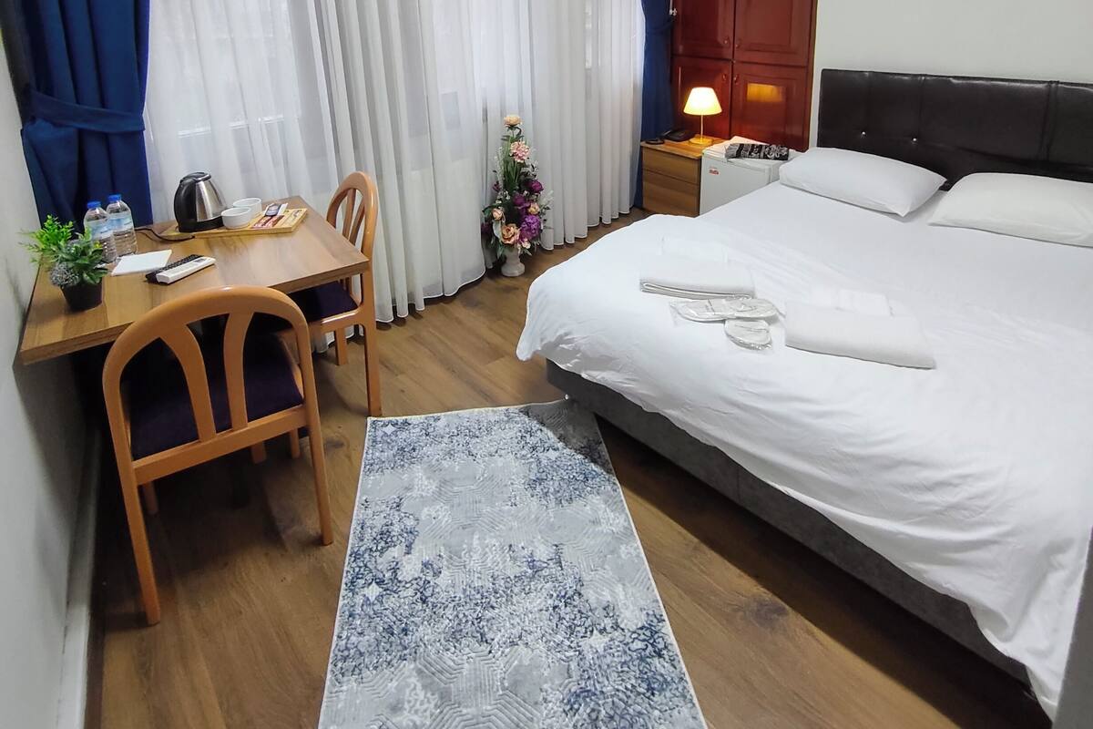 Quarto de hotel com cama, escrivaninha com cadeira, mesa de cabeceira, luminária, quadro na parede e janela com cortinas.