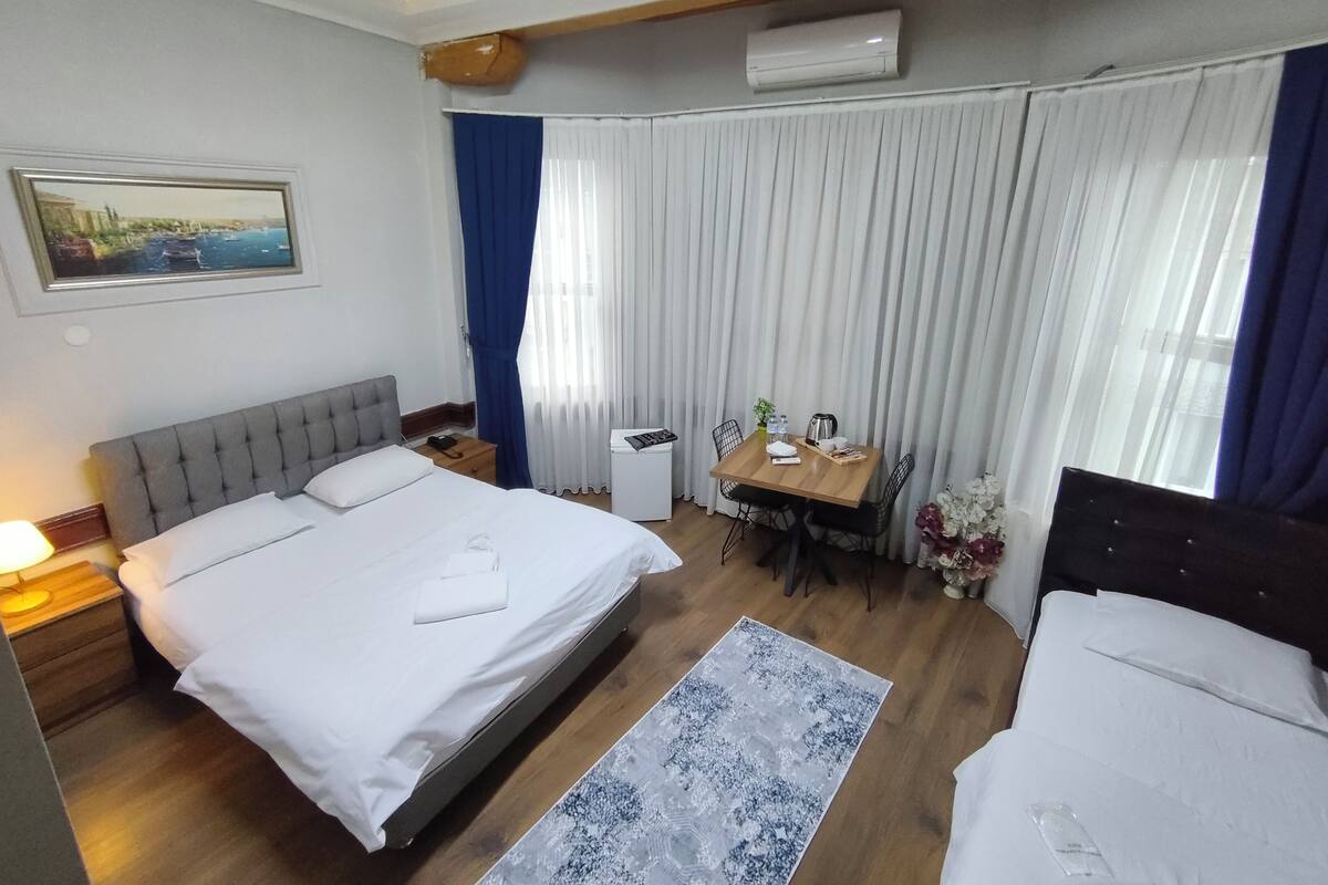Quarto de hotel com cama, mesa de cabeceira, luminária, aparelho de ar condicionado, janela com cortinas e uma pequena mesa com chaleira e flores.