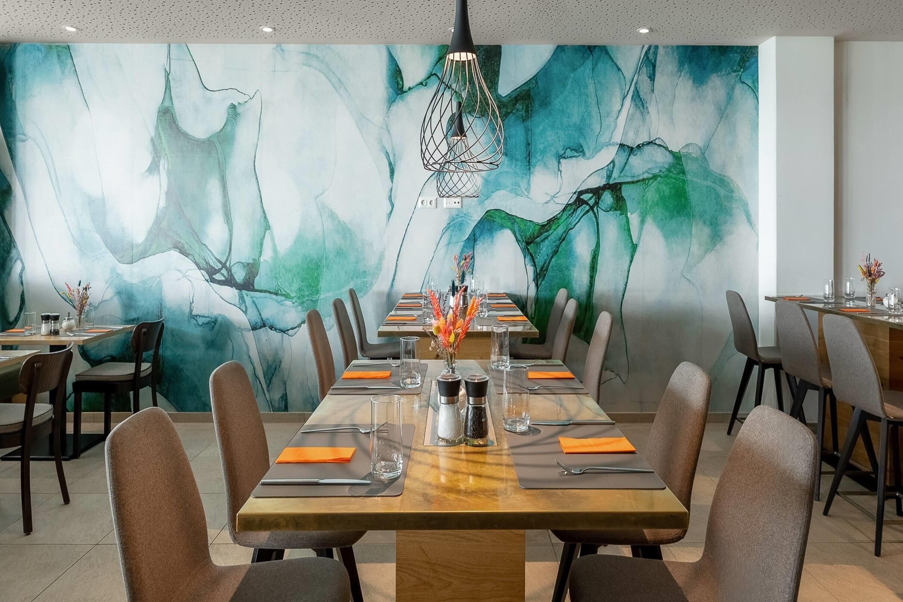 Uma área de jantar moderna com uma mesa longa posta para refeição, rodeada por cadeiras, e um grande mural abstrato na parede.