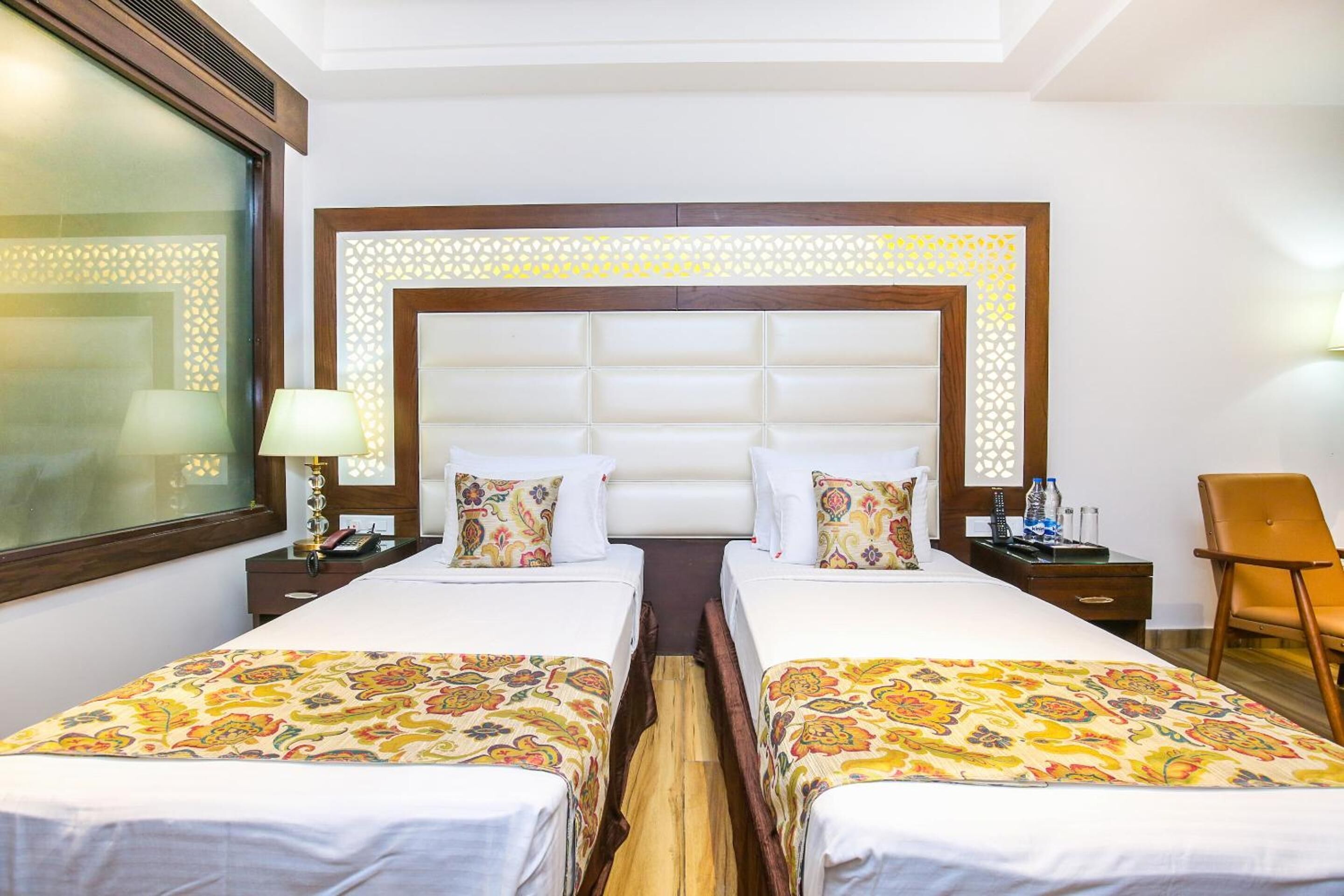 Quarto de hotel com duas camas, uma cabeceira de madeira, uma mesa de cabeceira com abajur, uma cadeira e uma janela com persianas.