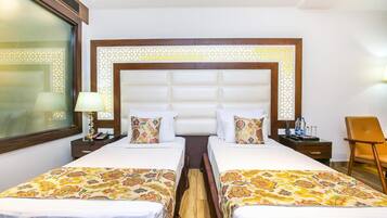 Quarto de hotel com duas camas, uma cabeceira de madeira, uma mesa de cabeceira com abajur, uma cadeira e uma janela com persianas.