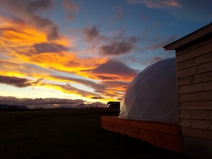 Uma cabana de madeira com uma tenda em forma de cúpula em frente, com o céu do pôr do sol e nuvens ao fundo.