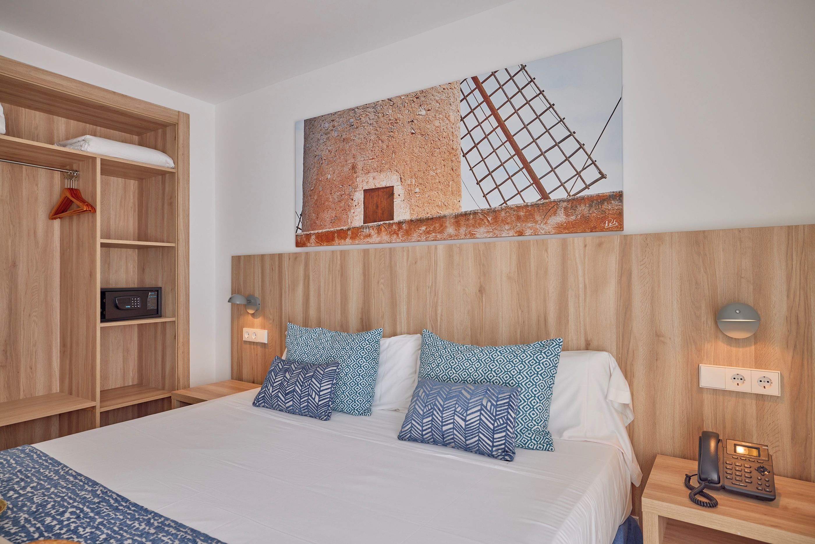 Quarto de hotel com cama, guarda-roupa de madeira, pintura, mesa de cabeceira com telefone e luminária fixada na parede.