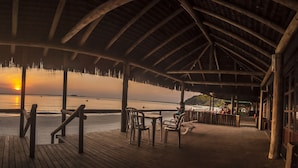 Um deck de madeira com telhado de palha dando vista para uma praia ao pôr do sol.
