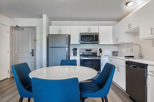 Uma cozinha moderna com armários brancos, uma geladeira de aço inoxidável, micro-ondas, forno e lava-louças. Há uma mesa branca redonda com cadeiras azuis.