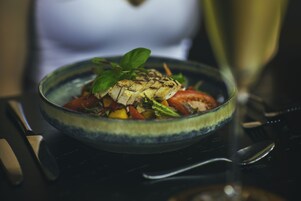 Uma tigela de comida com diversos vegetais e um pedaço de carne grelhada, acompanhada por garfo e faca sobre uma mesa.