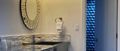 Um banheiro com bancada de granito, pia, espelho iluminado e porta que dá para um cômodo revestido com azulejos.