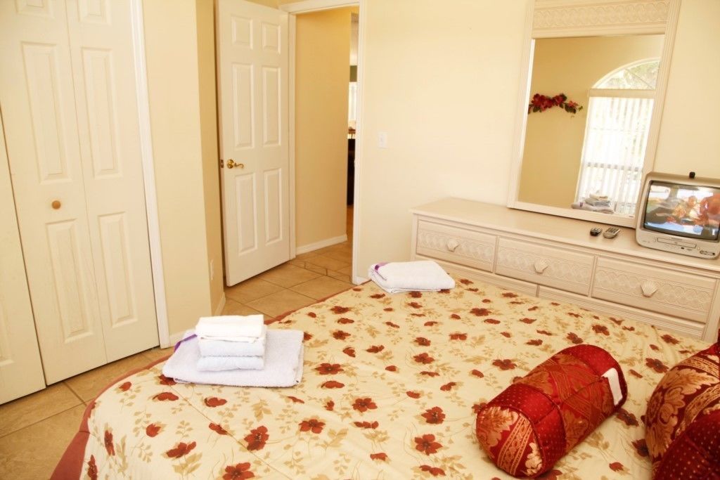 Quarto com colcha florida, penteadeira branca com espelho e porta que dá para outro cômodo.