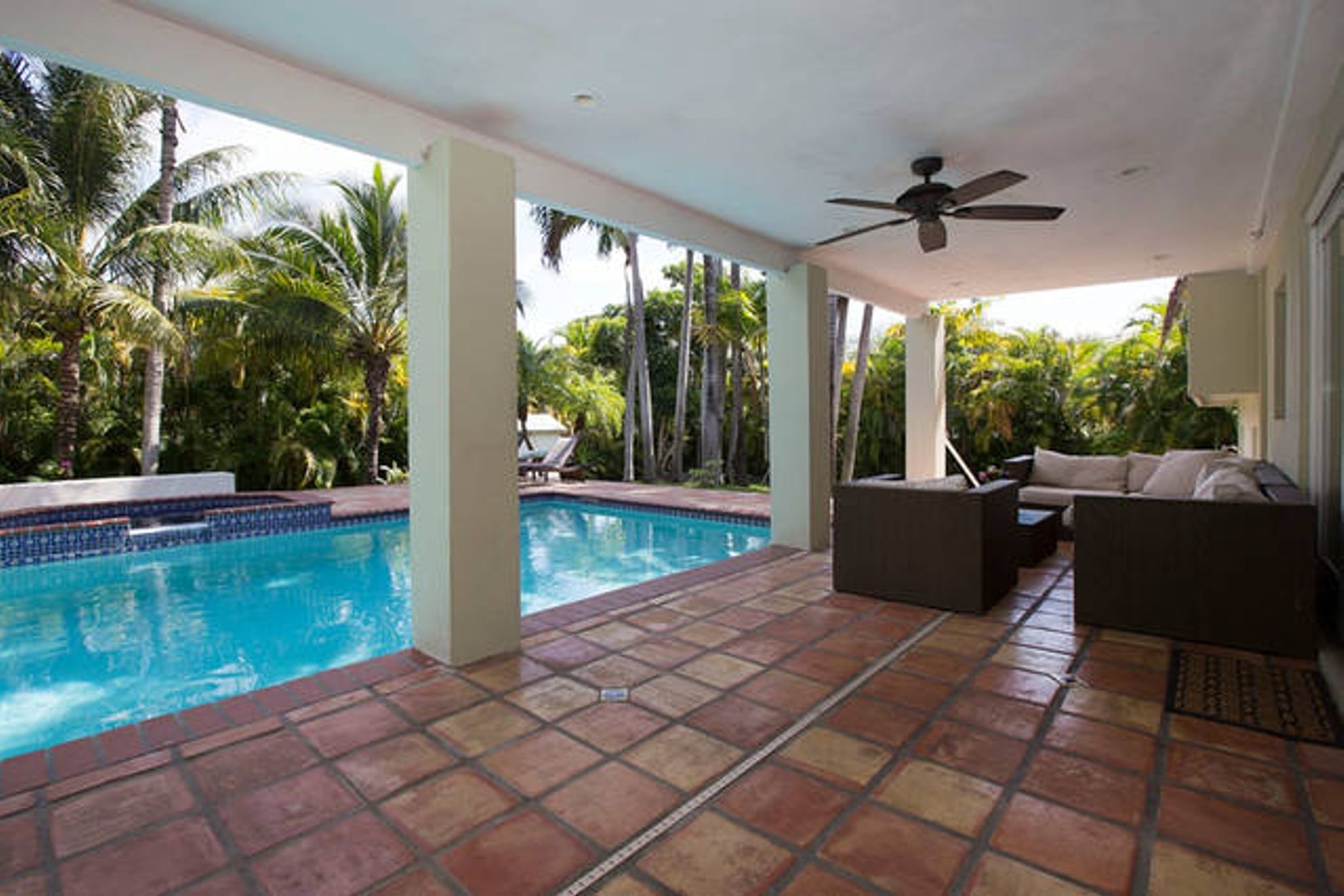 Uma área de piscina coberta com piso de azulejos, ventilador de teto e espaço de estar com sofá e cadeiras.
