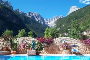 Piscina com muro de pedra e plantas em vasos, cercada por montanhas e casas.