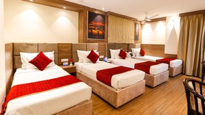 Quarto de hotel com quatro camas, cada uma com roupa de cama branca e detalhes vermelhos, cabeceiras de madeira e um quadro grande na parede.