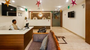 Uma área de recepção de hotel com um balcão, dois funcionários, um sofá e uma TV na parede.