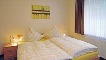 Uma cama de casal com edredom quadriculado amarelo e branco, luminária de cabeceira e mesa de cabeceira com gavetas.