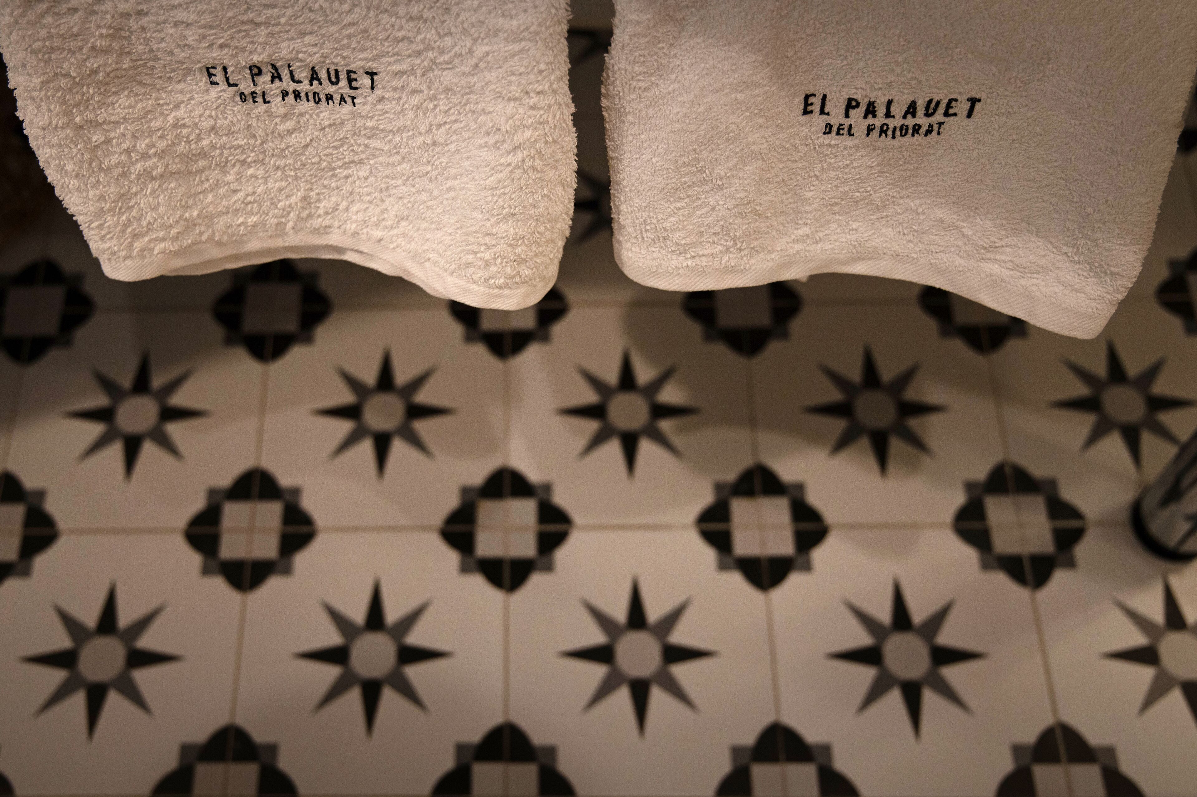 Duas toalhas brancas penduradas com o texto "EL PALAUET DEL PRIORAT" em um chão de azulejos com padrão de estrela.