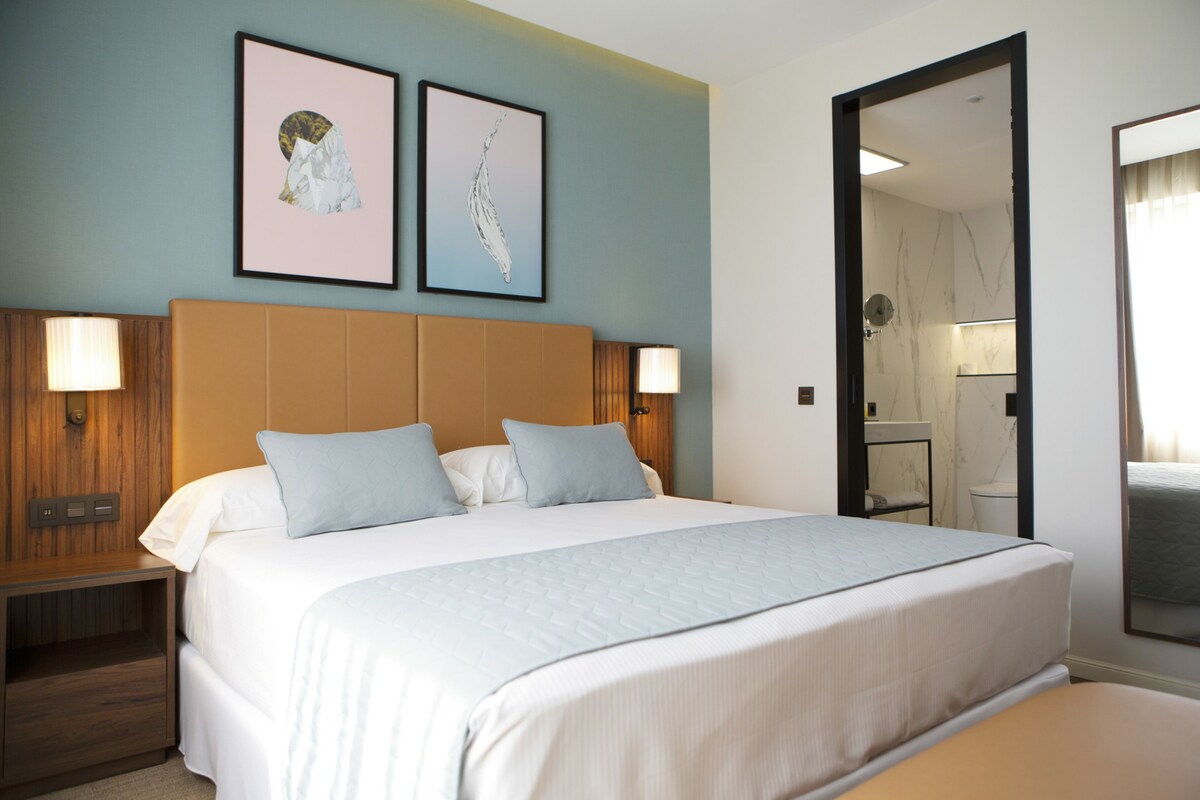 Quarto de hotel com uma cama grande, duas mesas de cabeceira, um criado-mudo, um espelho e obras de arte na parede.