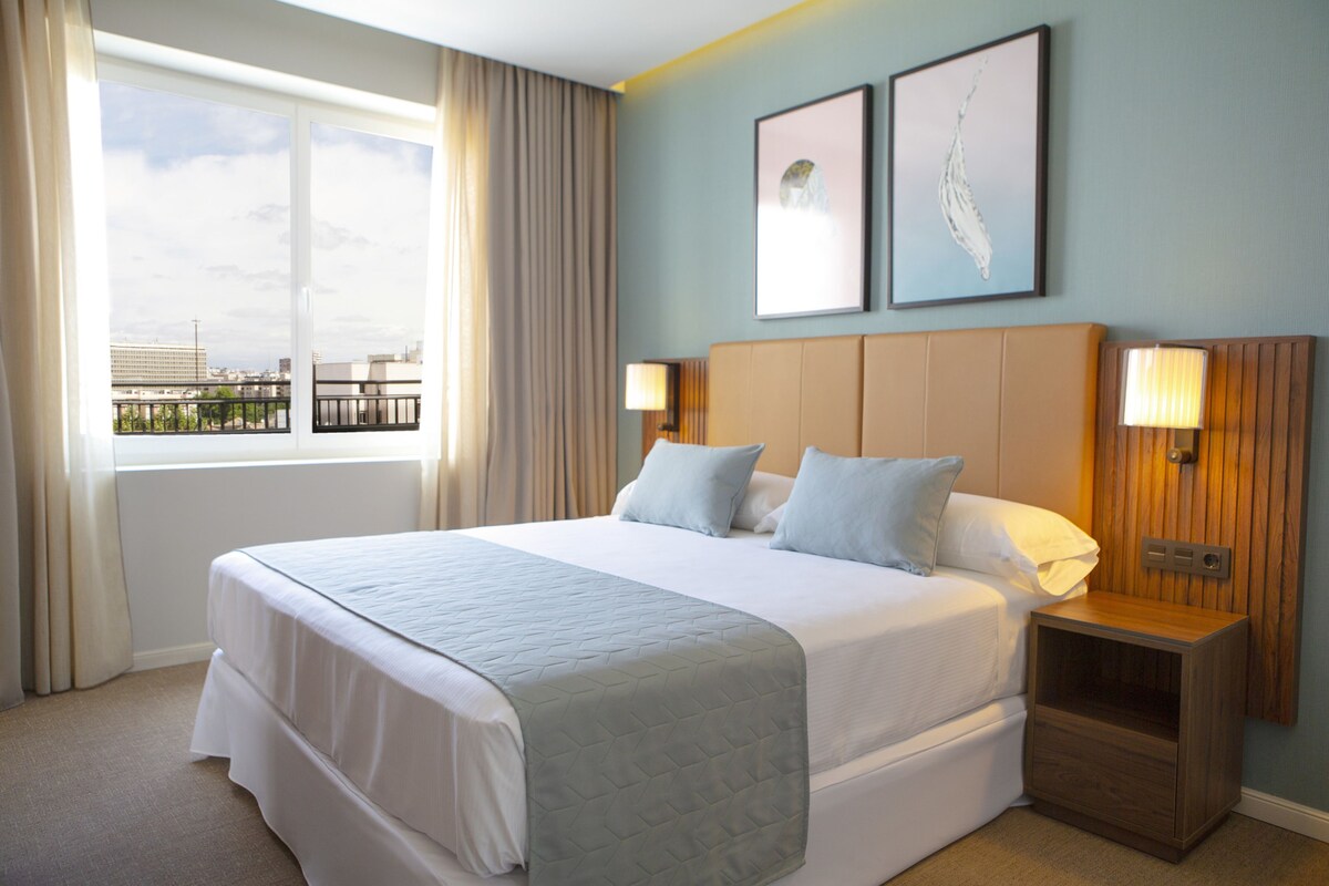 Quarto de hotel com uma cama grande, duas luminárias de cabeceira, uma mesa de cabeceira, uma janela com cortinas e duas obras de arte na parede.