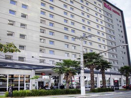 Um hotel de vários andares com fachada moderna, entrada coberta e palmeiras em primeiro plano.